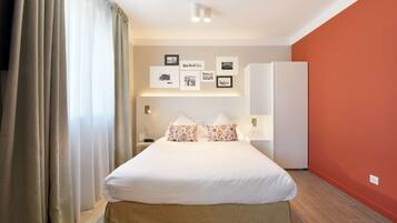 Quarto bem arrumado com cama, cortinas, prateleira na parede com quadros e guarda-roupa branco.