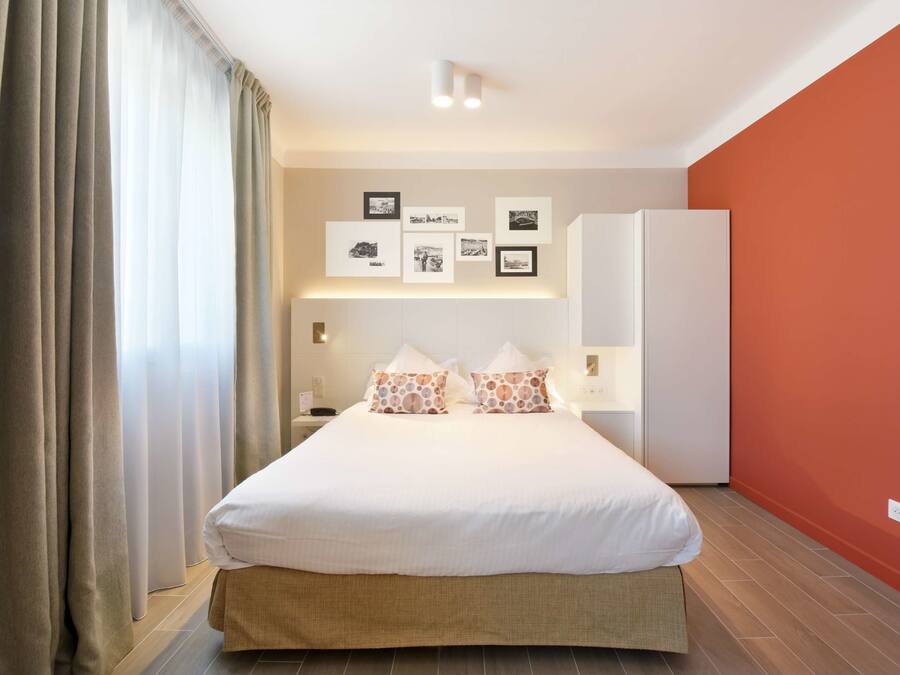 Quarto bem arrumado com cama, cortinas, prateleira na parede com quadros e guarda-roupa branco.