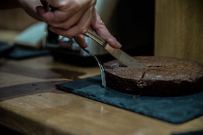 Uma mão cortando um bolo redondo com uma faca em uma tábua de madeira.