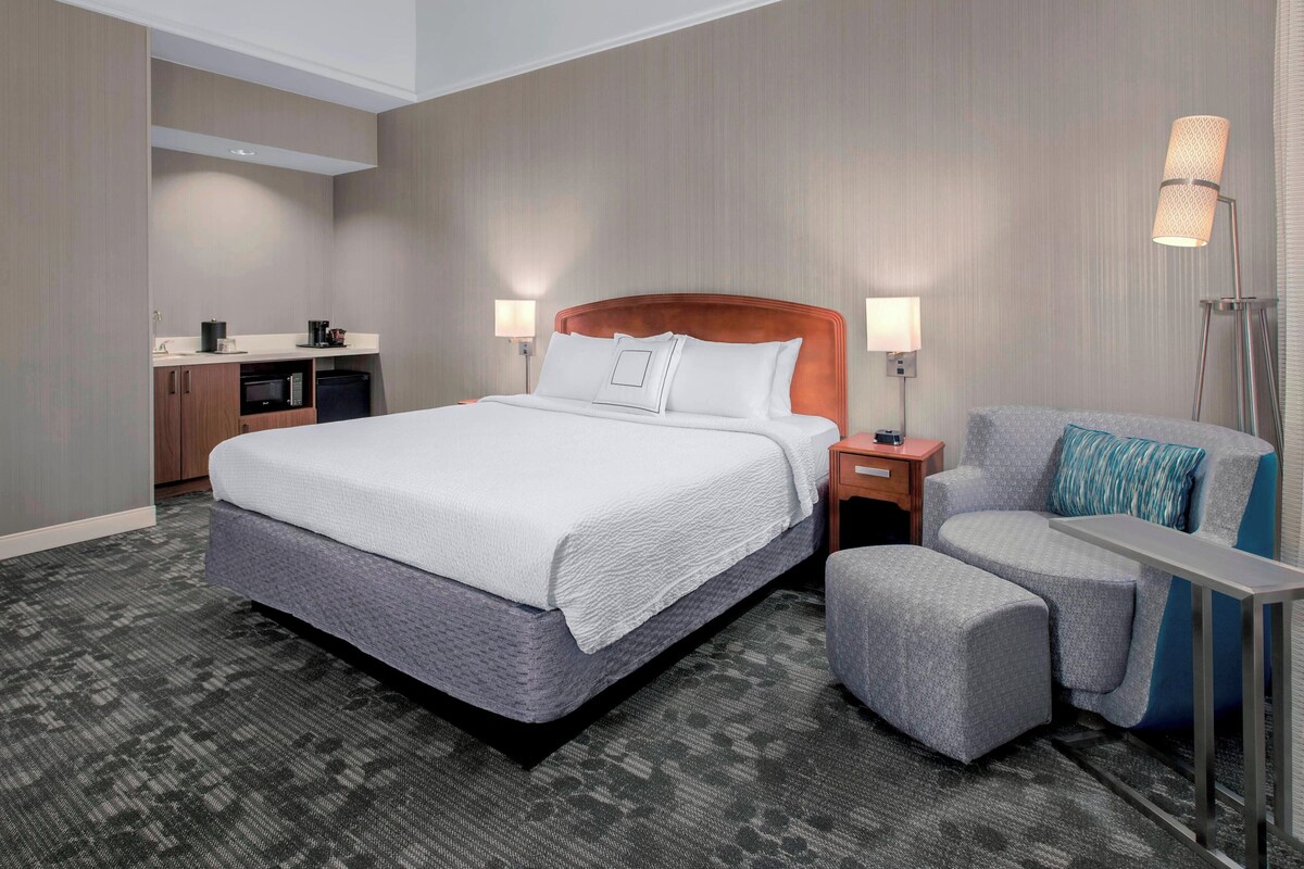 Quarto de hotel com uma cama grande, uma escrivaninha, uma cadeira e um abajur.