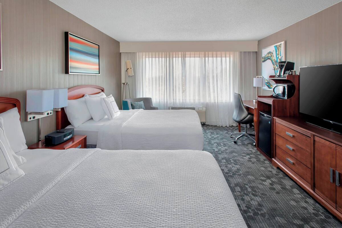 Quarto de hotel com duas camas, televisão, escrivaninha e janela com cortinas.