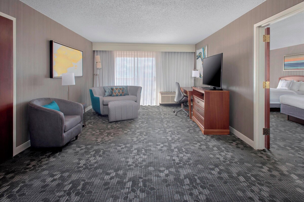 Quarto de hotel com um sofá cinzento, uma poltrona cinzenta, uma escrivaninha com televisão e uma cama.