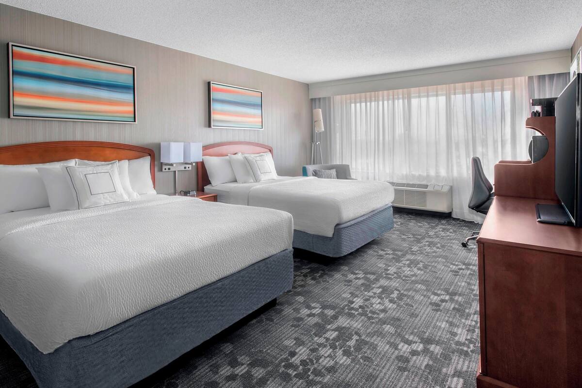 Quarto de hotel com duas camas, uma escrivaninha, uma cadeira, uma televisão e amplas janelas com cortinas.