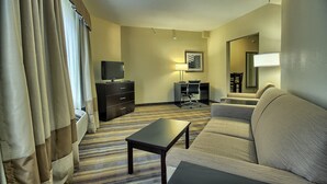 Quarto de hotel com sofá, mesa pequena, televisão, escrivaninha com cadeira e luminária.