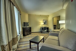 Quarto de hotel com sofá, mesa pequena, televisão, escrivaninha com cadeira e luminária.