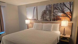 Quarto de hotel com uma cama grande, duas luminárias de cabeceira, um criado-mudo e uma parede com três quadros emoldurados.