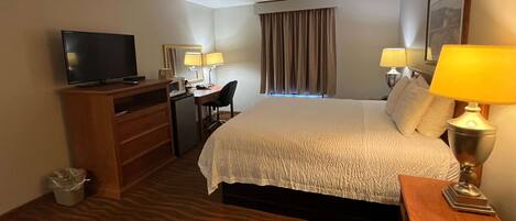 Quarto de hotel com cama, escrivaninha com cadeira, televisão e duas luminárias.