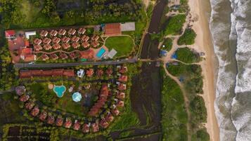 Vista aérea de um resort com vários edifícios tipo bangalô, uma piscina central e uma praia adjacente à propriedade.