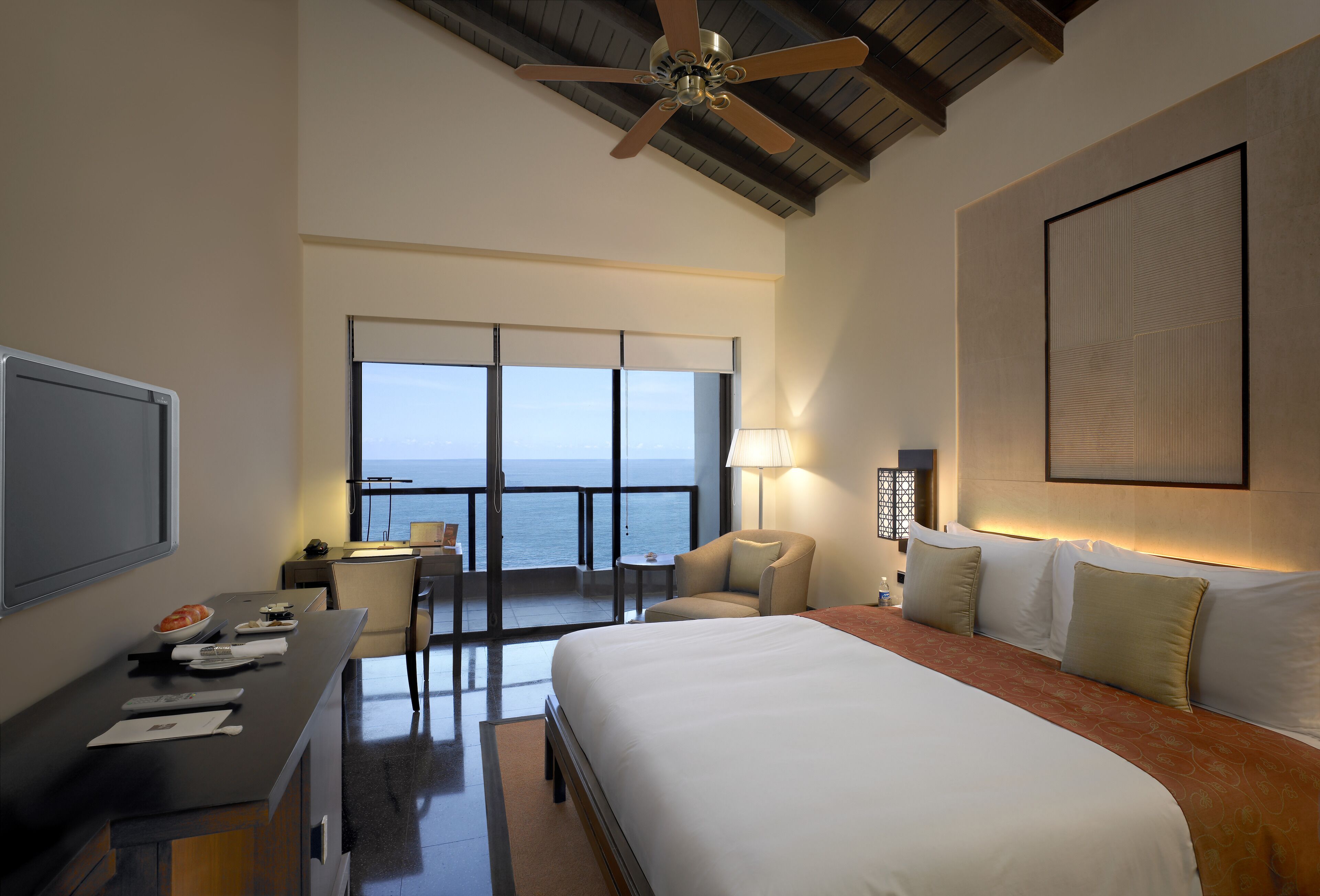 Quarto de hotel com uma cama grande, uma escrivaninha com cadeira, uma mesinha, uma luminária e vista para o mar por grandes janelas.