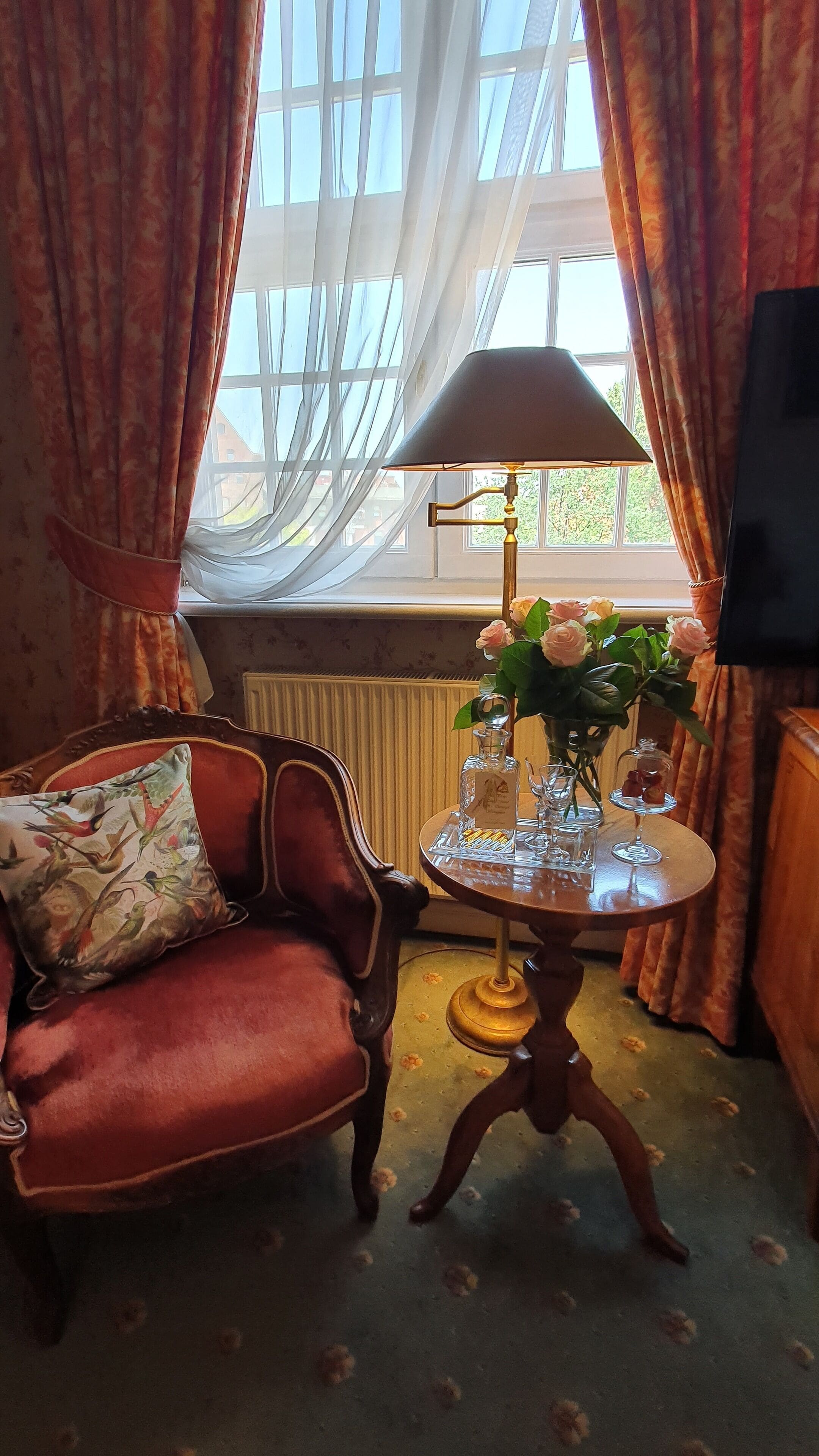 Quarto com sofá vermelho, mesa com abajur, vaso com flores e janela com cortinas.