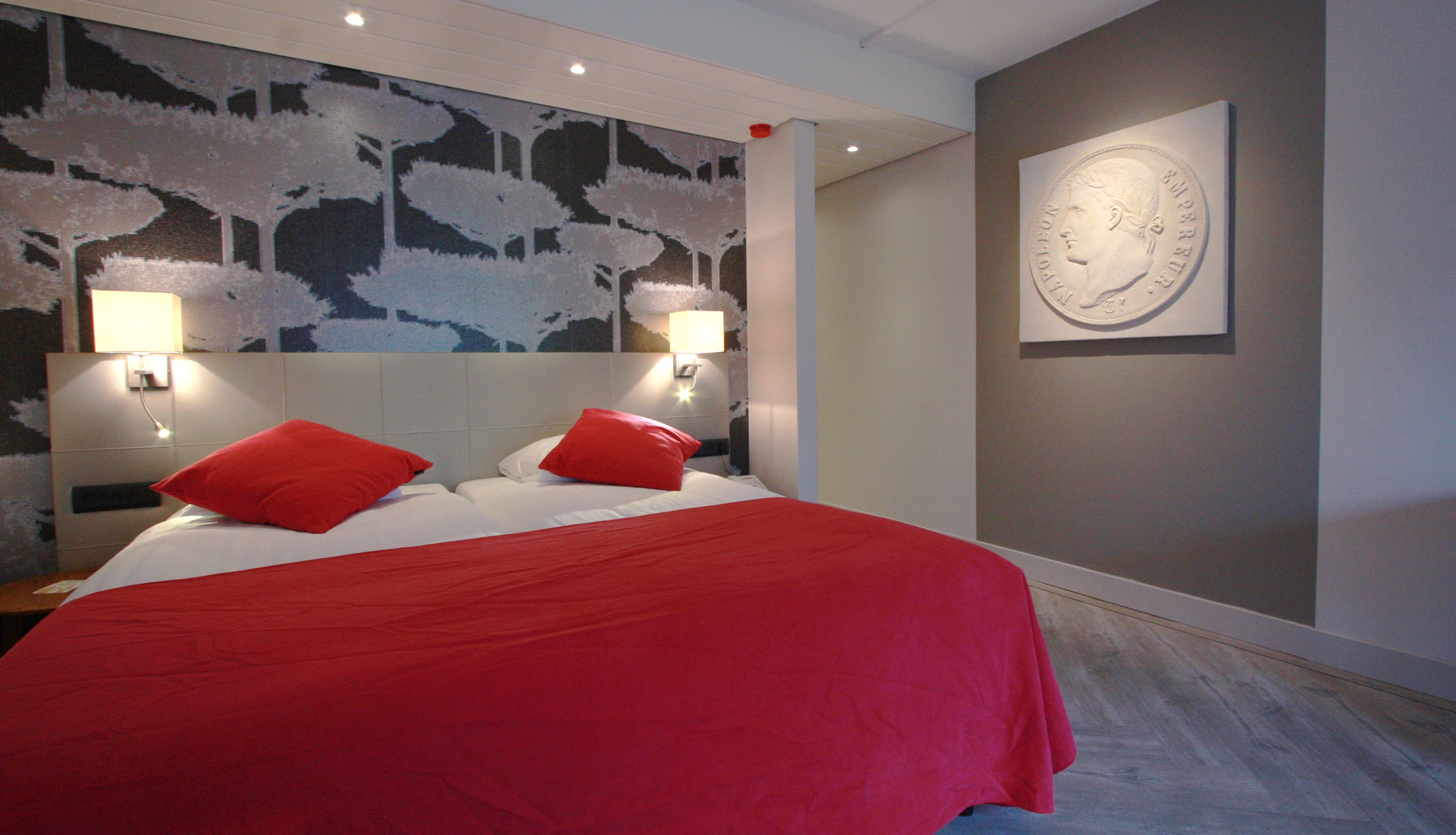 Quarto de hotel moderno com uma cama grande, roupa de cama vermelha, duas luminárias fixadas na parede e uma obra de arte emoldurada.