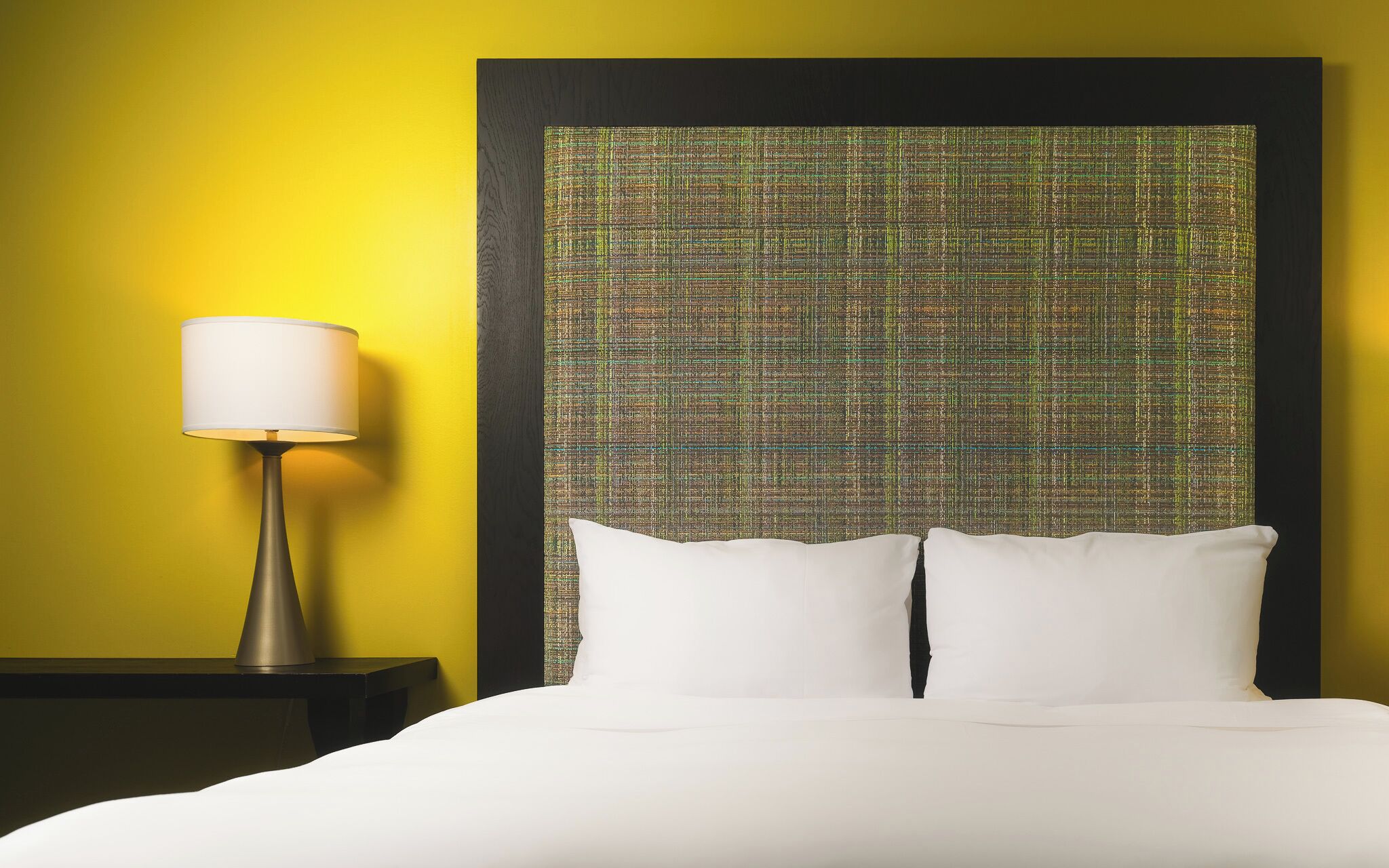 Quarto de hotel com cama, almofadas brancas, mesa de cabeceira e abajur.