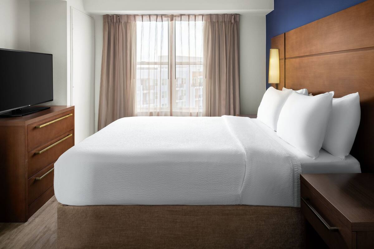 Quarto de hotel com uma cama grande, uma TV sobre uma cômoda de madeira e uma janela com cortinas.