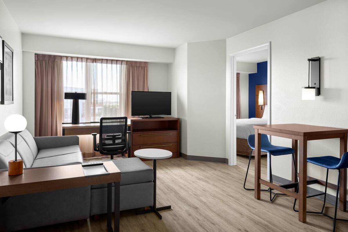 Quarto de hotel moderno com sofá, escrivaninha com computador, televisão e área de refeições com cadeiras.