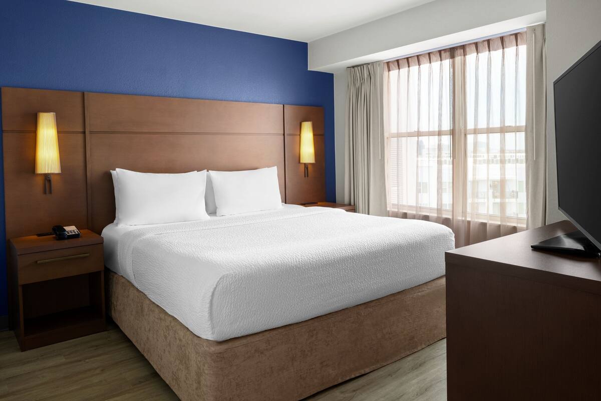 Quarto de hotel com uma cama grande, duas luminárias de cabeceira, uma escrivaninha com televisão e uma janela com cortinas.