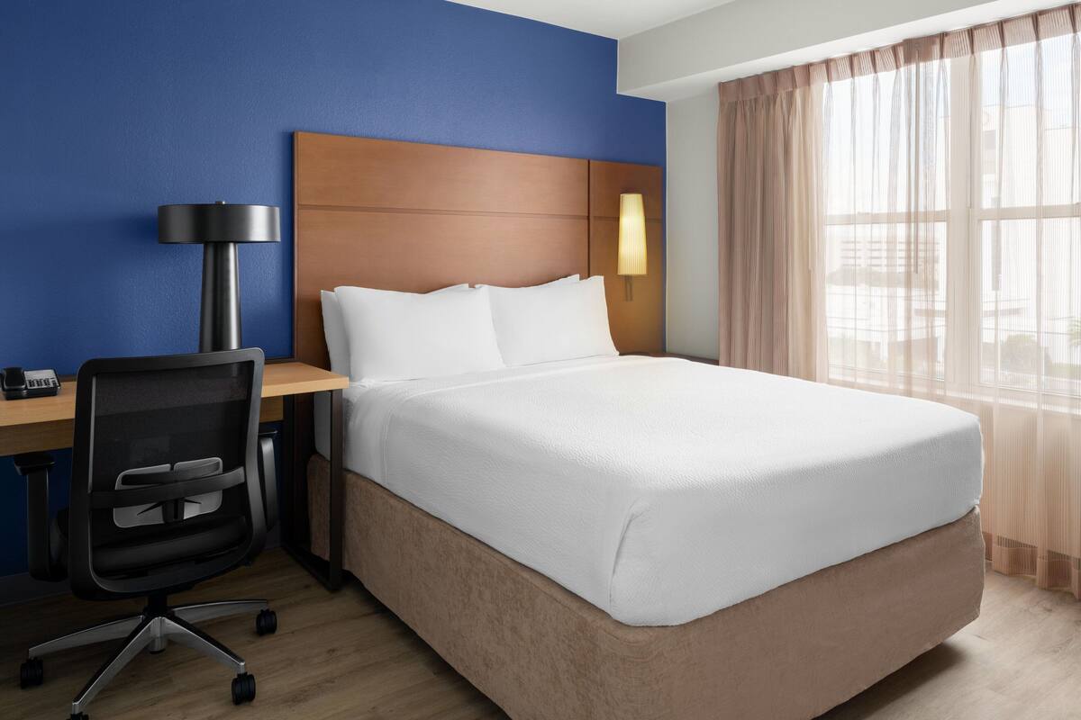 Quarto de hotel com uma cama grande, uma escrivaninha com cadeira, um abajur e uma janela com cortinas leves.