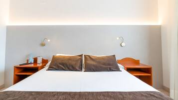 Cama bem arrumada com duas almofadas, mesa de cabeceira com abajur, e criado-mudo com telefone e controle remoto.