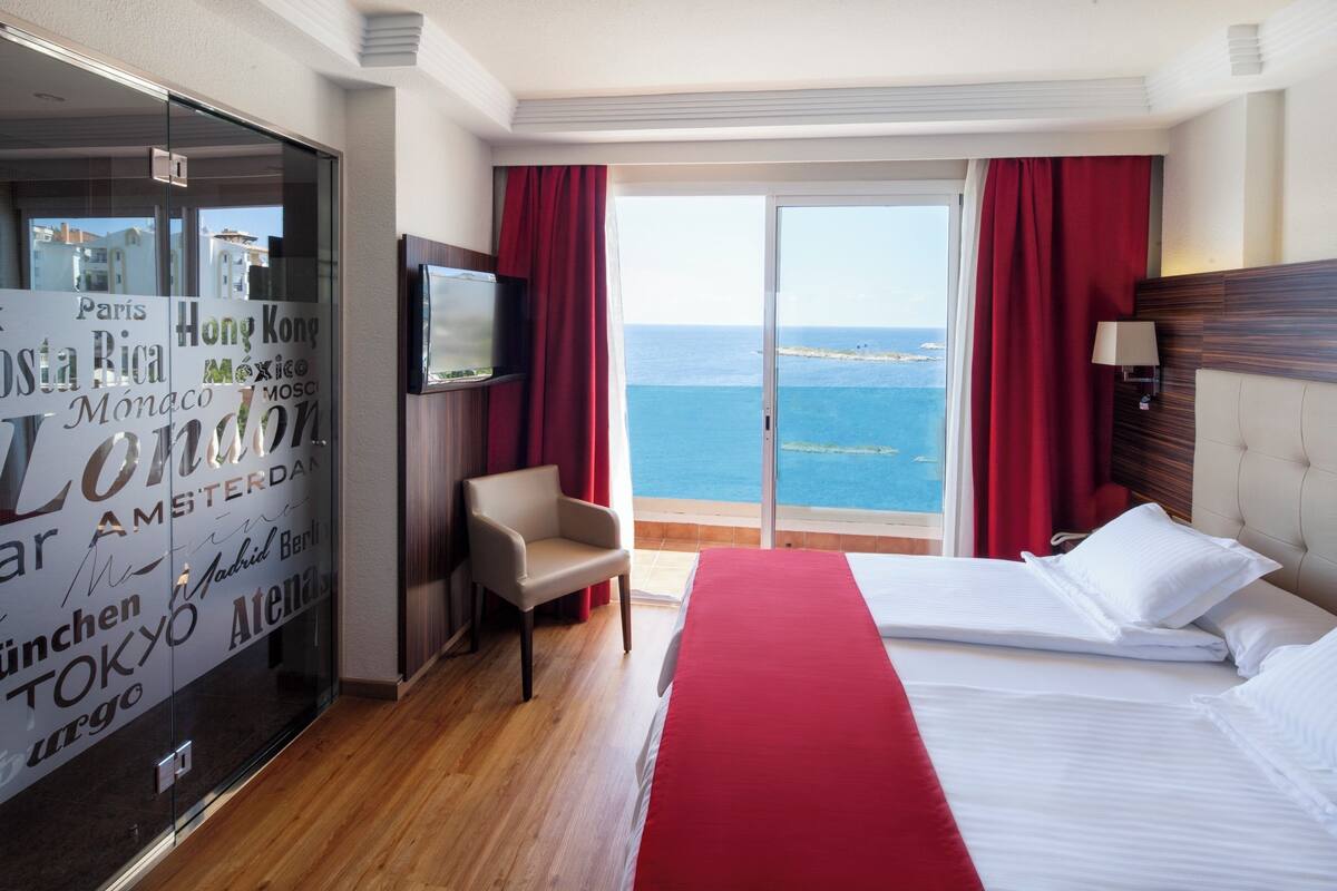 Quarto de hotel com cama, poltrona, televisão e uma janela ampla com vista para o mar.