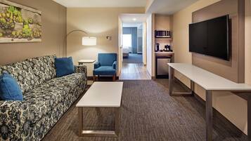 Quarto de hotel com sofá estampado, uma mesa pequena, televisão e área de cozinha compacta.