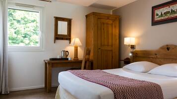 Quarto de hotel com uma cama, um guarda-roupa de madeira, uma escrivaninha com abajur e uma janela com cortinas.