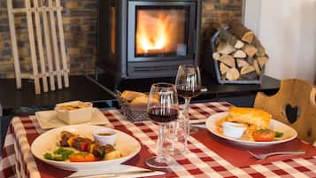 Uma mesa de jantar com toalha quadriculada, pratos de comida, copos de vinho e uma lareira ao fundo.
