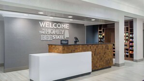 Uma área de recepção de hotel moderna, com um balcão de madeira, uma placa de boas-vindas grande e um quiosque digital.
