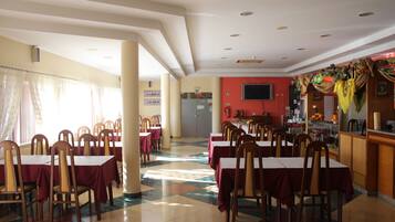 Restaurante com mesas postas para refeições, área de bar e balcão com exposição de frutas e legumes.