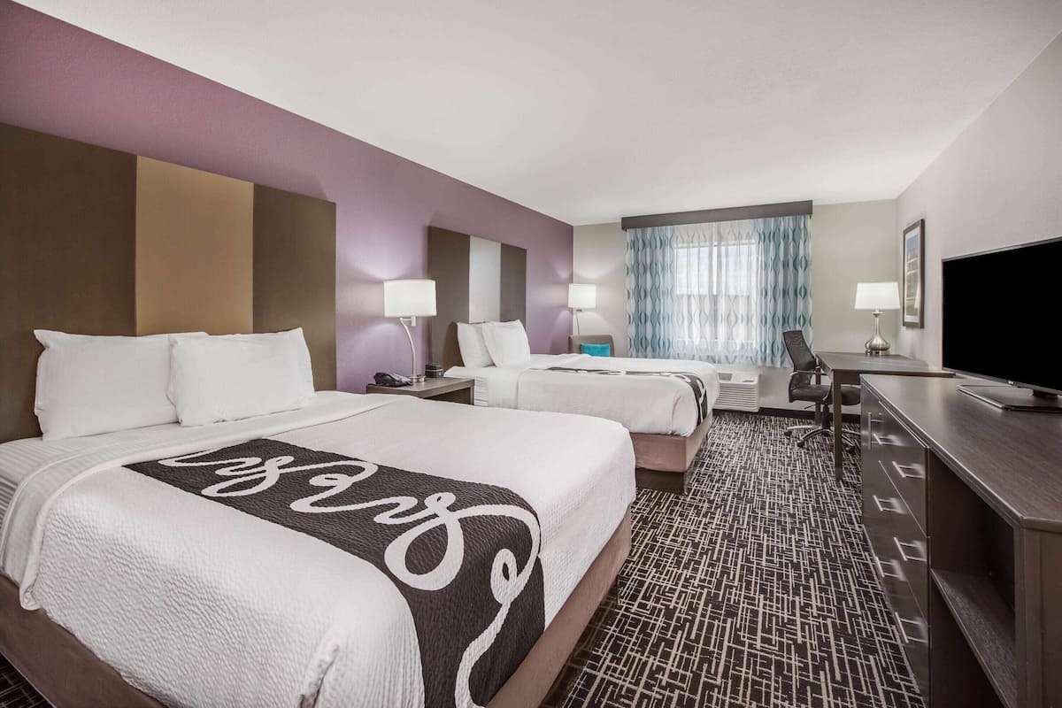 Quarto de hotel com duas camas, uma escrivaninha, uma televisão e um tapete estampado.