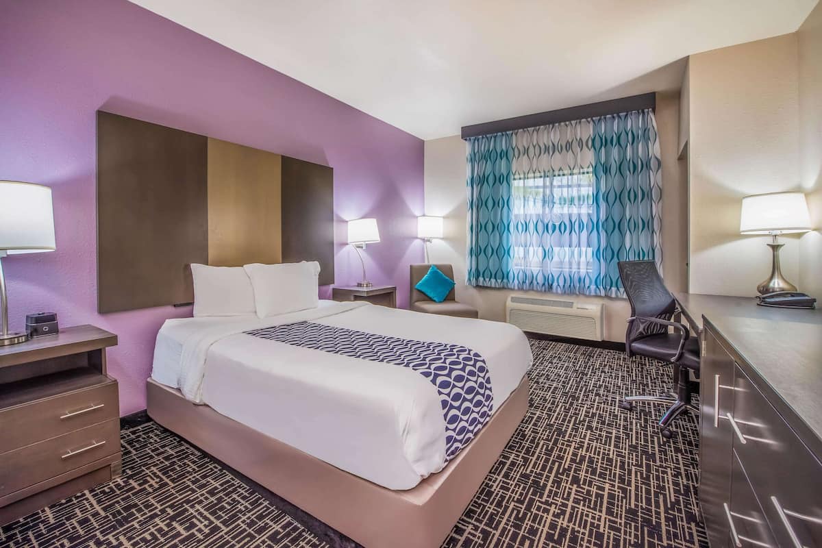 Quarto de hotel com uma cama grande, duas mesas de cabeceira com luminárias, uma escrivaninha com cadeira e uma janela com cortinas estampadas.
