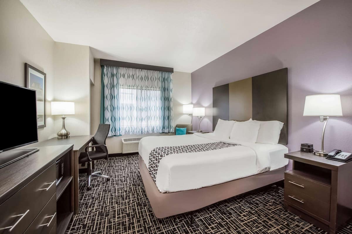 Quarto de hotel com cama, escrivaninha, cadeira, televisão e luminárias.