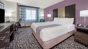 Quarto de hotel com uma cama grande, uma escrivaninha com gavetas, uma cadeira, uma luminária, um telefone e uma janela com cortinas estampadas.