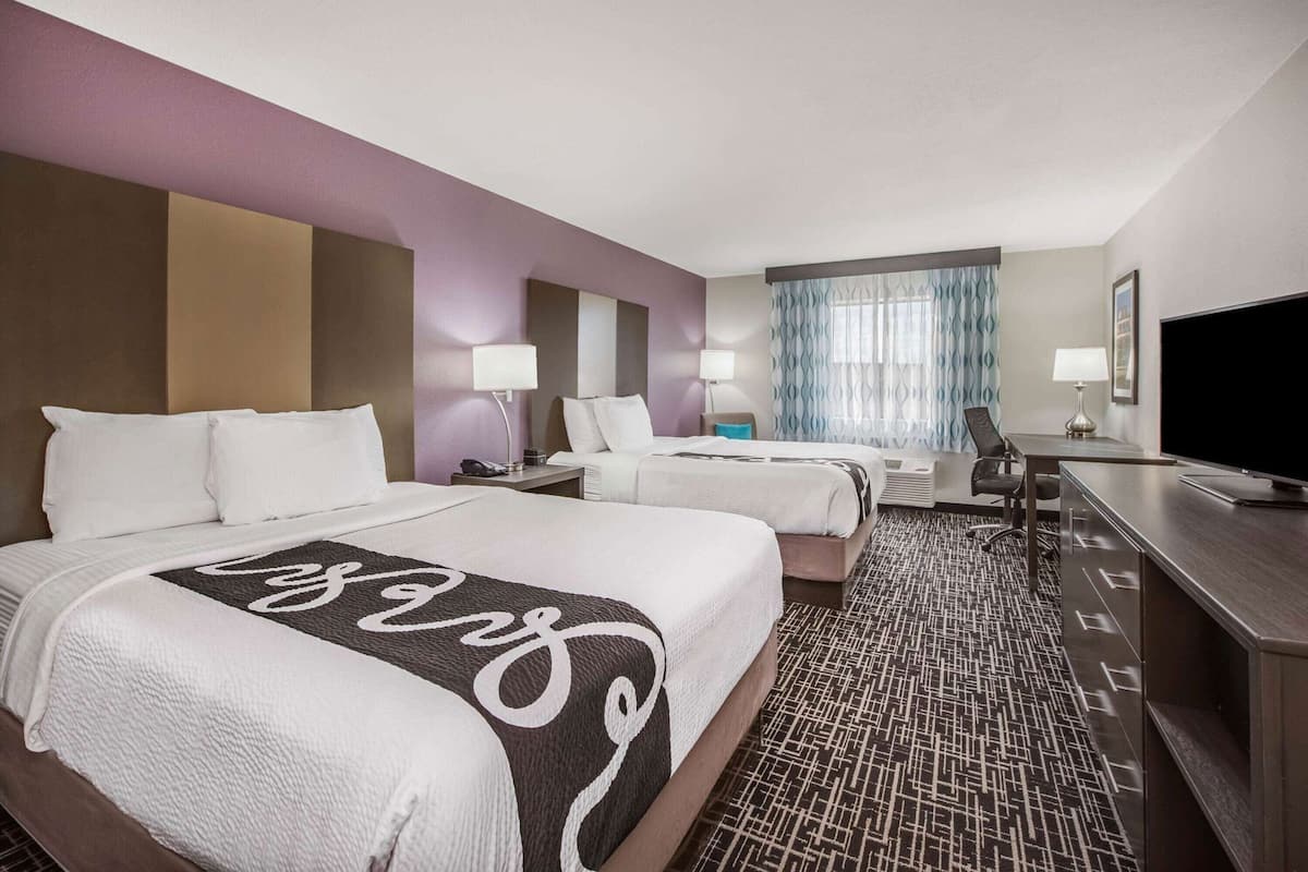 Quarto de hotel com duas camas, uma escrivaninha, uma cadeira, uma televisão e um tapete estampado.