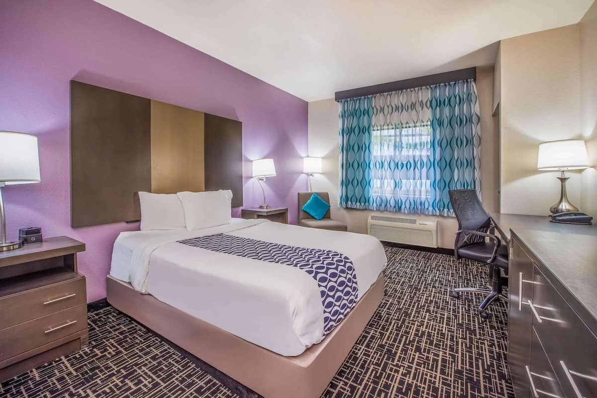 Quarto de hotel com cama, mesas de cabeceira, escrivaninha, cadeira e janela com cortinas.