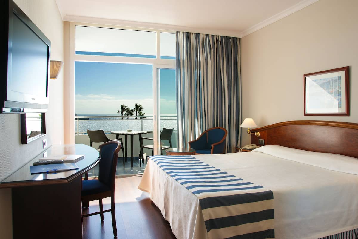 Quarto de hotel com uma cama grande, uma escrivaninha com computador, uma cadeira, televisão e vista para o mar.