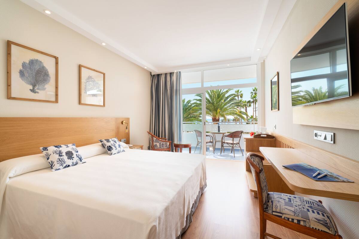 Quarto de hotel com uma cama grande, escrivaninha, cadeiras, televisão e varanda com palmeiras.