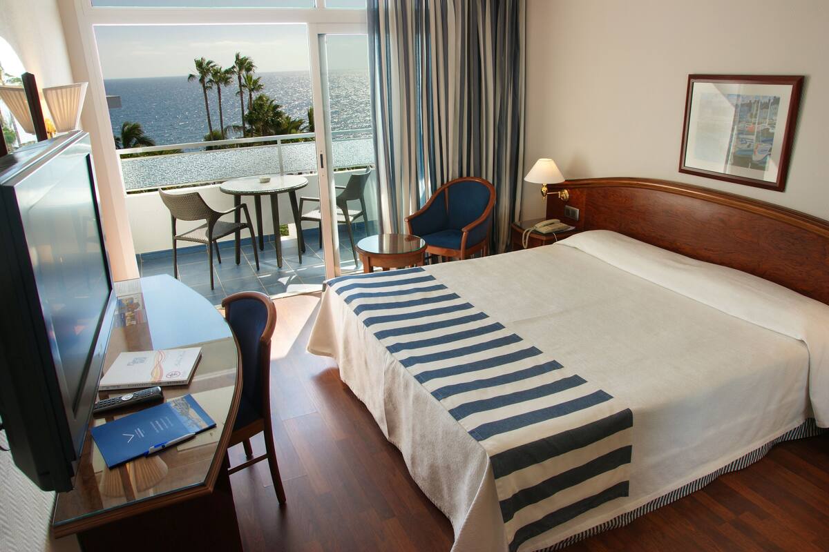 Quarto de hotel com uma cama, uma escrivaninha com um laptop, uma cadeira, uma mesa pequena e vista para o oceano.