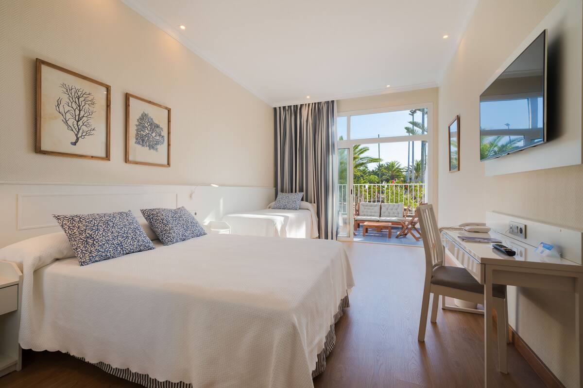 Quarto de hotel com uma cama grande, uma escrivaninha, uma cadeira, uma televisão e varanda com vista para palmeiras.