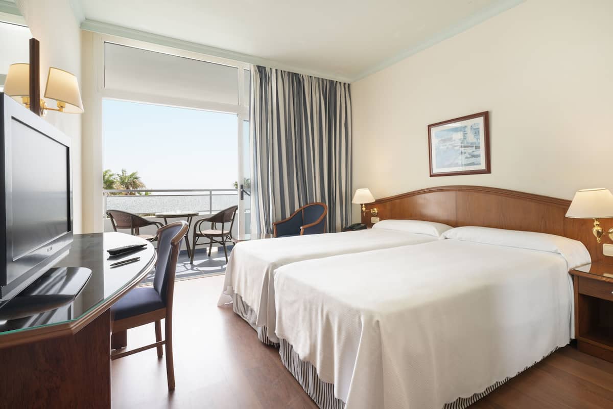 Quarto de hotel com duas camas, uma escrivaninha com televisão, uma cadeira, varanda com vista e um quadro pendurado na parede.