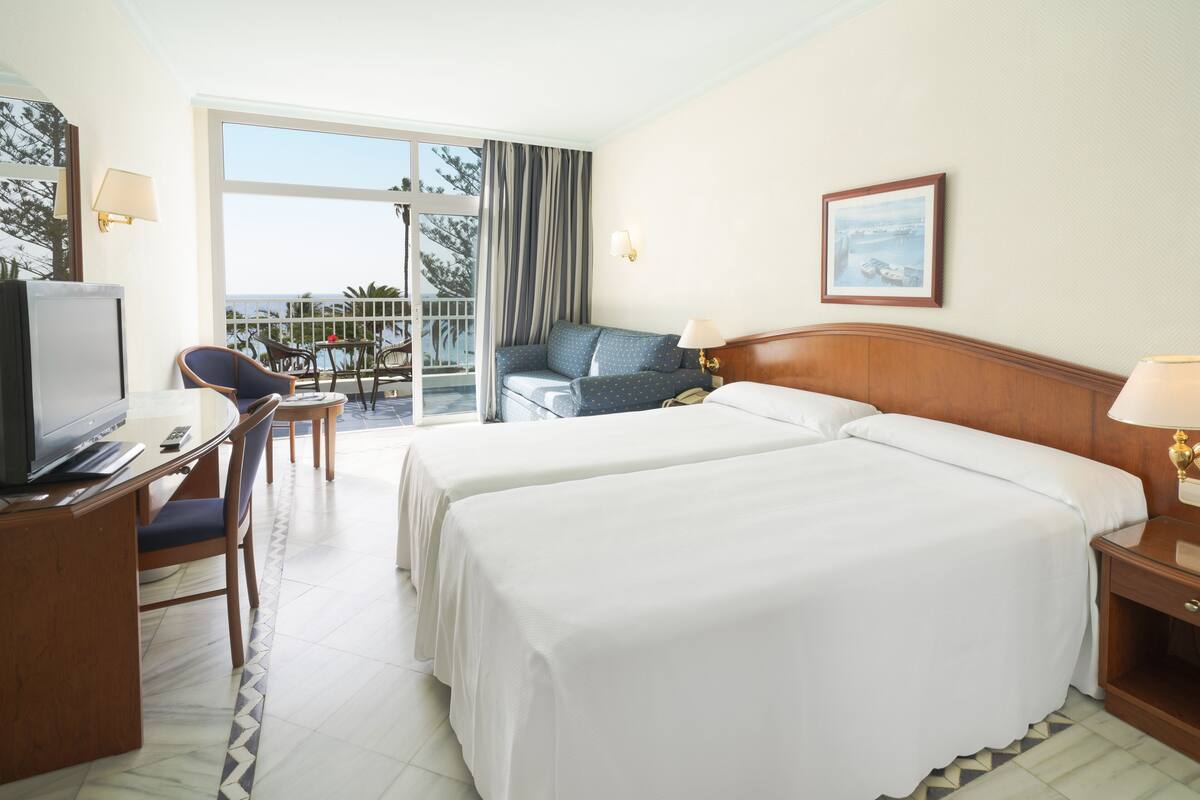 Quarto de hotel com duas camas, uma mesa de trabalho com televisão, uma cadeira, varanda com vista e um quadro na parede.