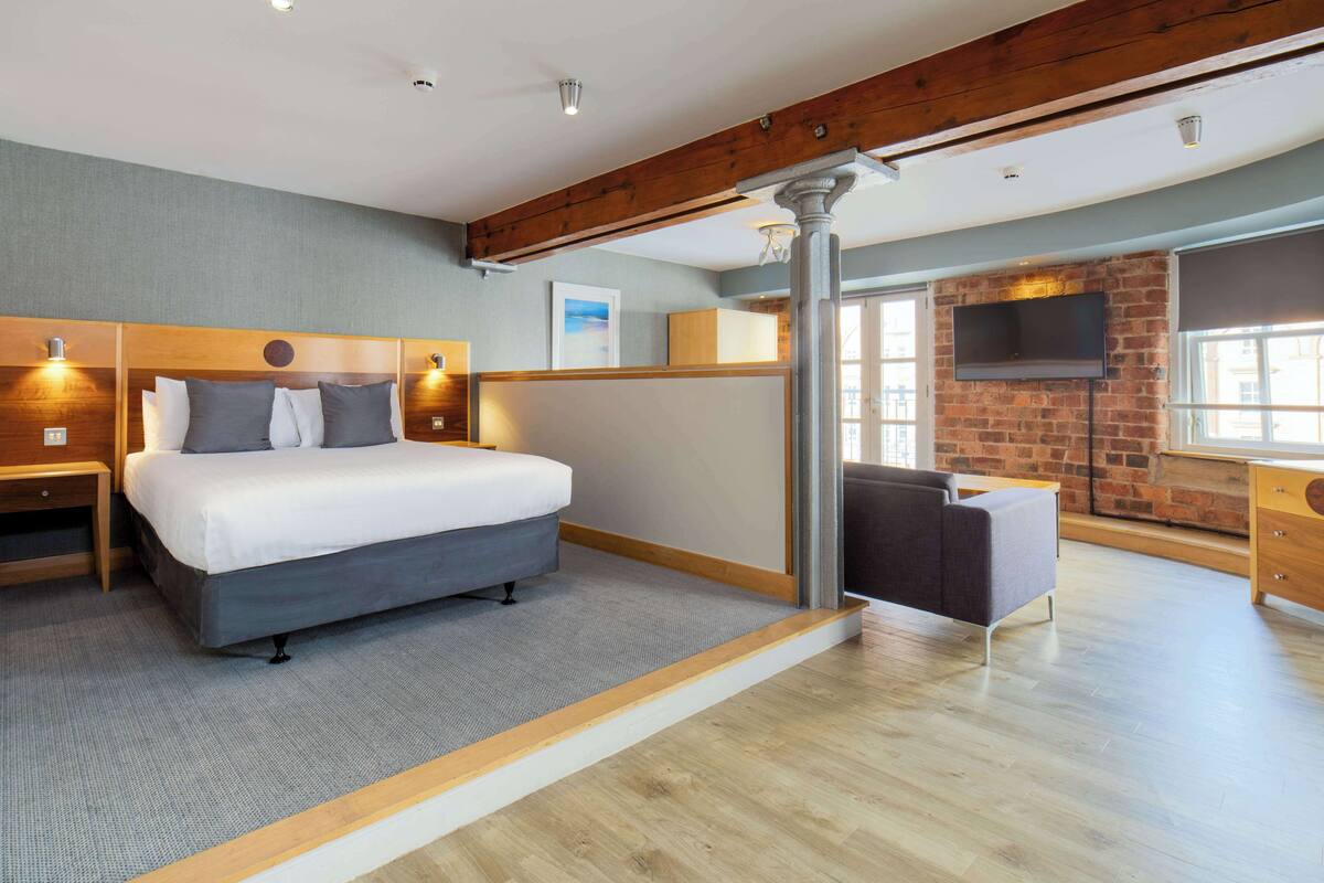 Quarto de hotel moderno com cama grande, escrivaninha, cadeira, televisão e parede de tijolos.
