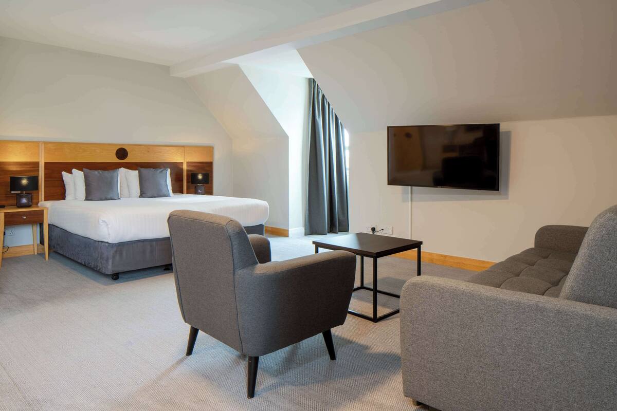 Quarto de hotel moderno com cama, escrivaninha, televisão e dois poltronas.