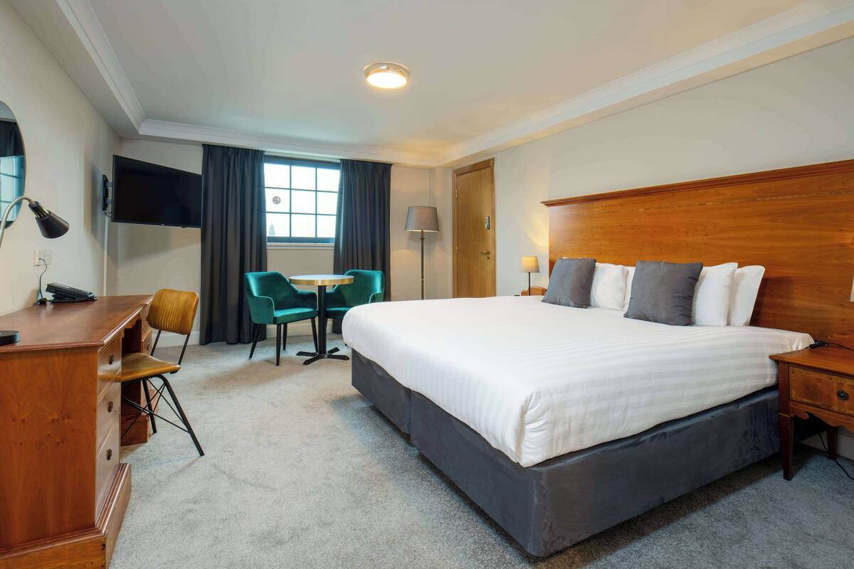 Quarto de hotel com uma cama grande, uma escrivaninha, uma cadeira, uma televisão e uma janela com cortinas.