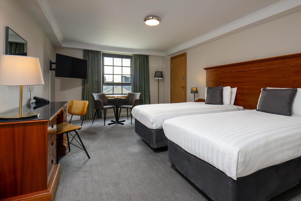 Quarto de hotel com duas camas, uma escrivaninha com cadeira, televisão e janela com cortinas.