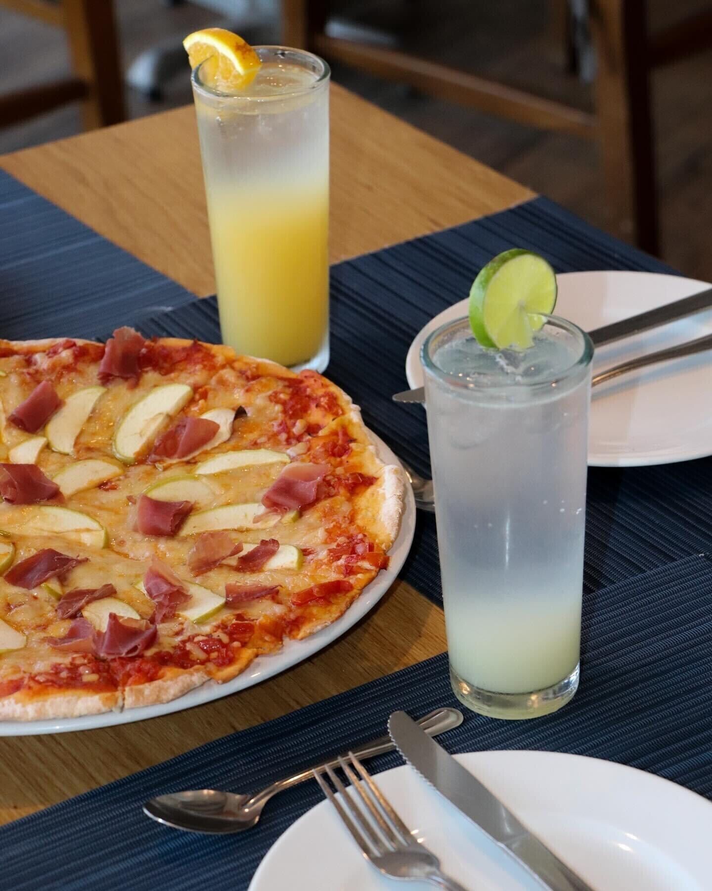 Pizza com fiambre e fatias de maçã, um copo de bebida gelada com uma fatia de limão, e um garfo e uma faca sobre uma mesa.