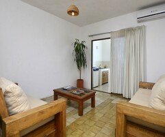 Sala de estar com duas cadeiras de madeira, uma mesa de centro, uma planta em vaso e vista para uma cozinha através de uma porta de vidro.