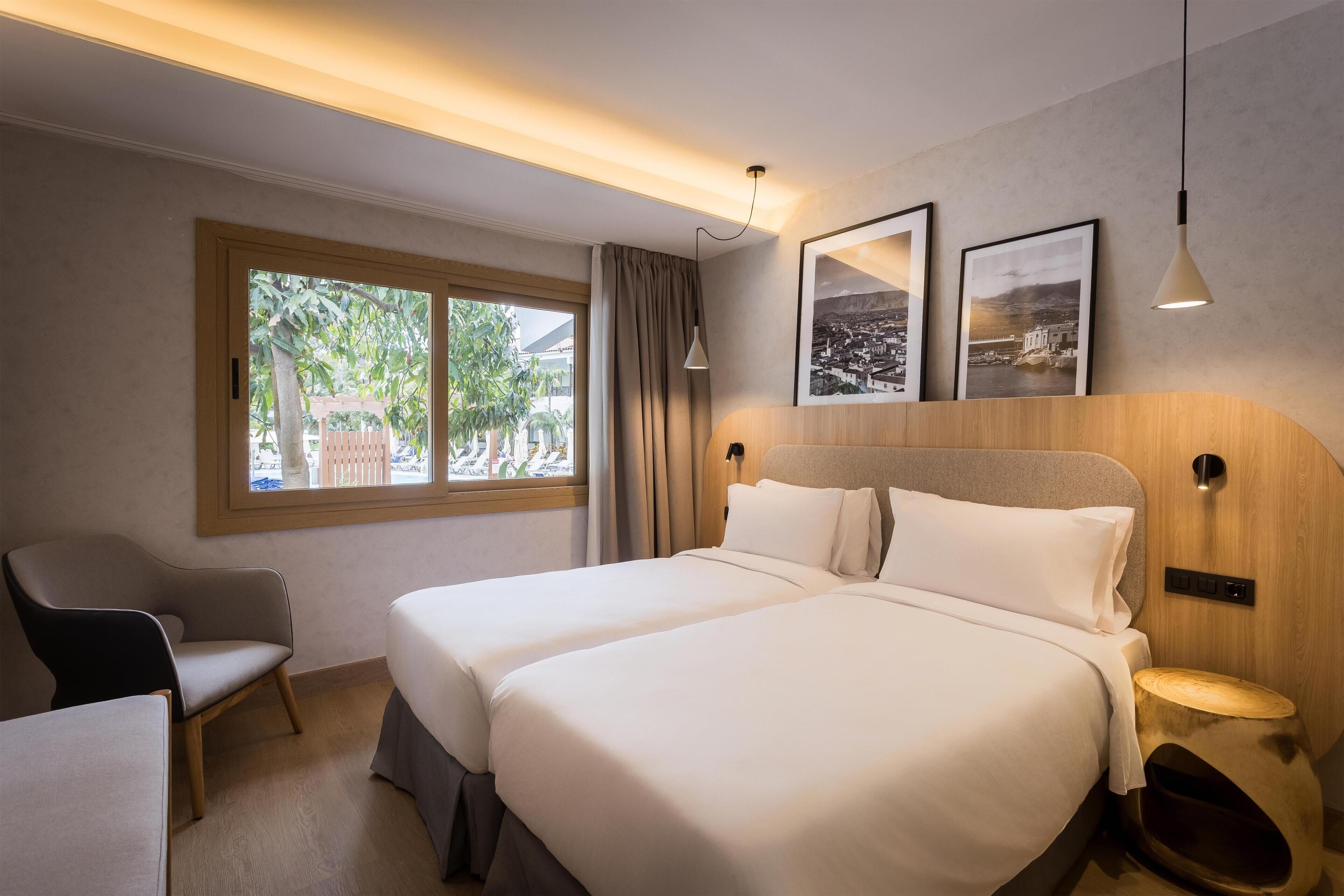 Quarto de hotel com duas camas, uma escrivaninha, uma cadeira, uma janela com vista e dois quadros na parede.