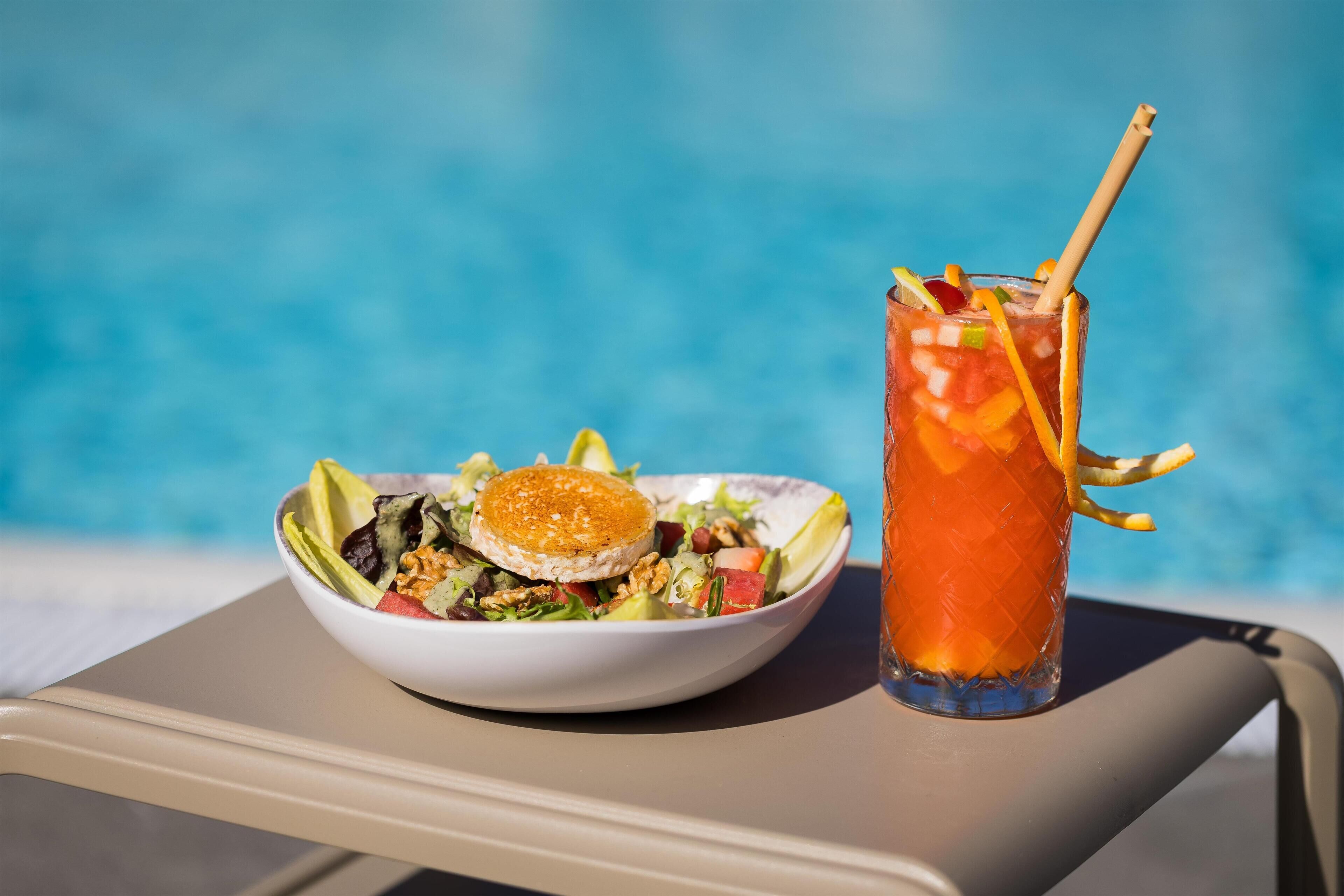 Uma salada com um hambúrguer e um copo alto de chá gelado com canudo, colocados sobre uma mesa perto de uma piscina.