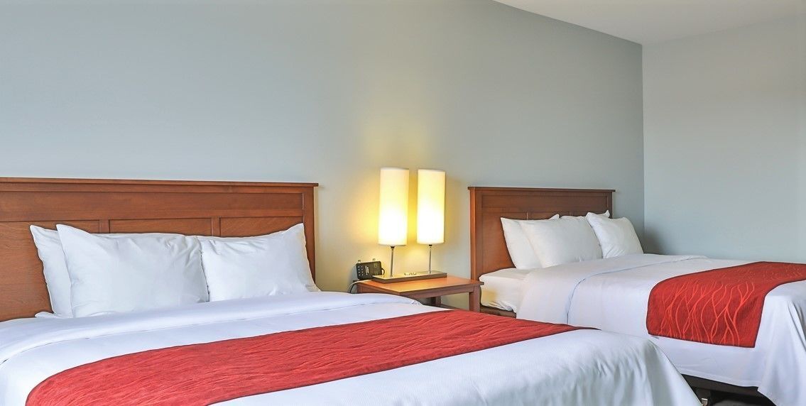 Quarto de hotel com duas camas, cada uma com roupa de cama branca e detalhes vermelhos, uma mesa de cabeceira com abajur, e um candeeiro fixo na parede.