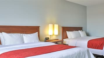 Quarto de hotel com duas camas, cada uma com roupa de cama branca e detalhes vermelhos, uma mesa de cabeceira com abajur, e um candeeiro fixo na parede.