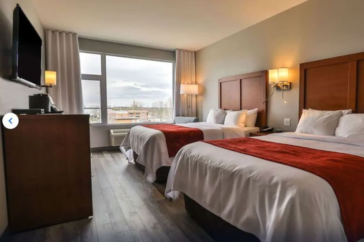 Quarto de hotel com duas camas, televisão e janela com vista para edifícios.